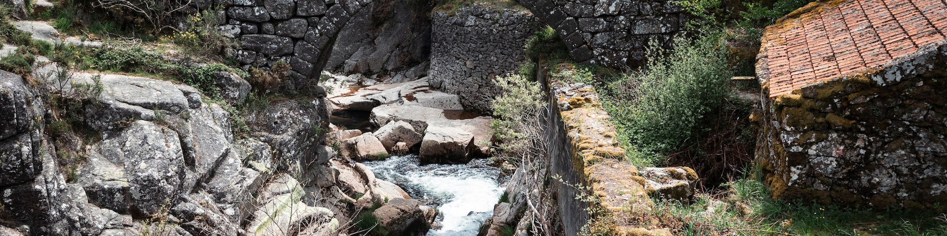 Uma ponte de pedra sobre um rio corrente, rodeada por terreno rochoso e vegetação.