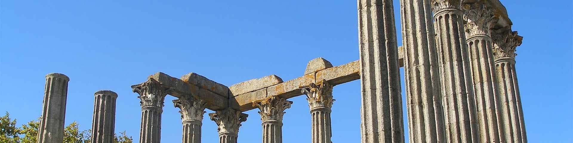 Colunas de pedra antiga com esculturas detalhadas, ao fundo um céu azul límpido.