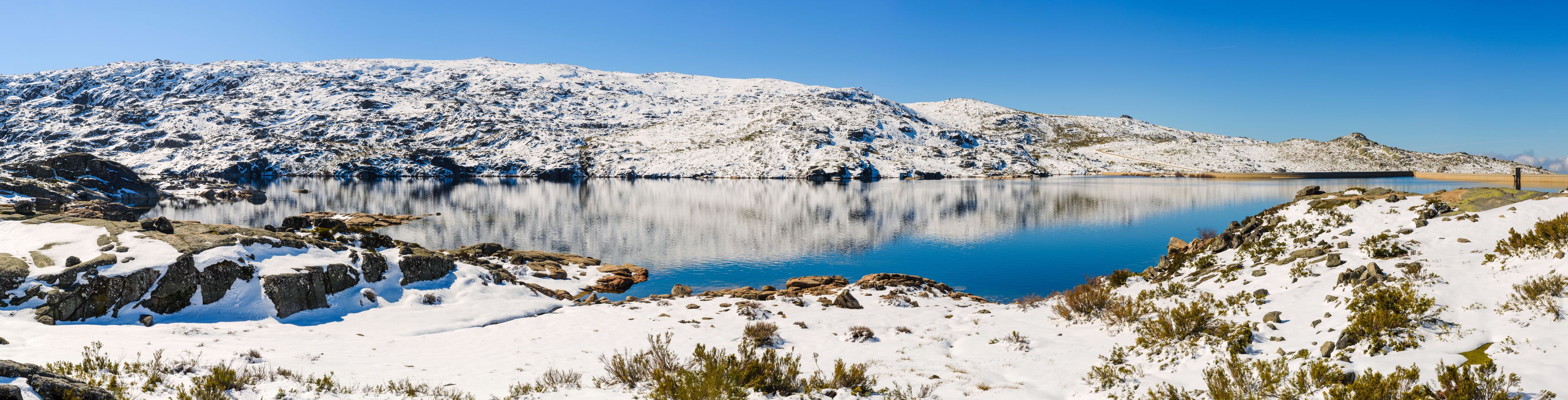 Cordilheira coberta de neve, um lago calmo e terreno rochoso.