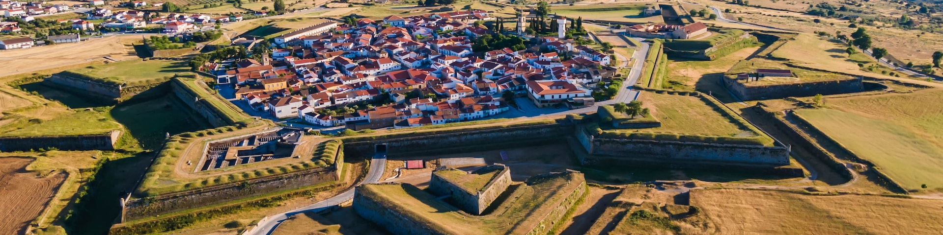 Vista aérea de uma cidade histórica cercada por fortificações e colinas ondulantes.