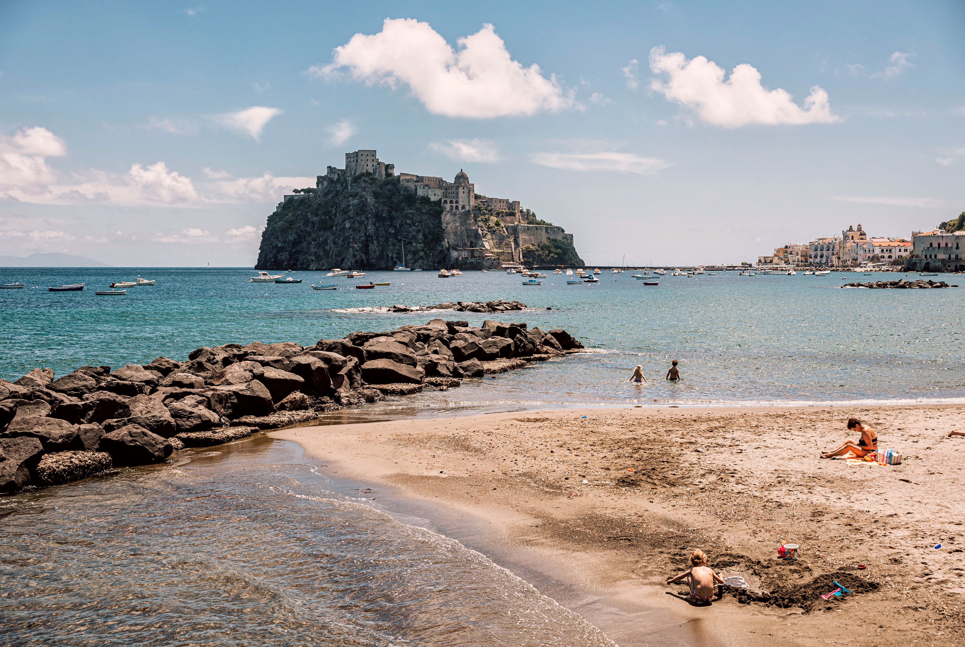 Uma praia costeira com paredões rochosos, mar calmo e edifícios históricos ao fundo.