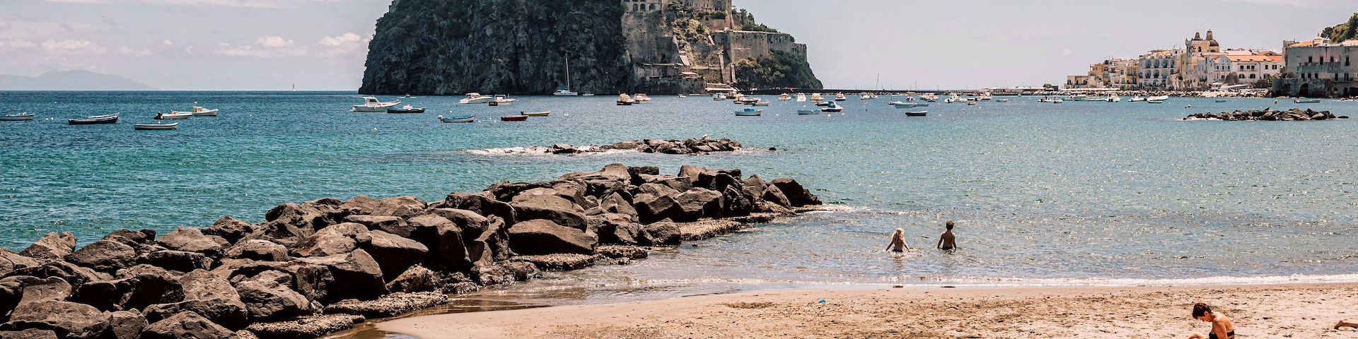 Uma praia costeira com paredões rochosos, mar calmo e edifícios históricos ao fundo.