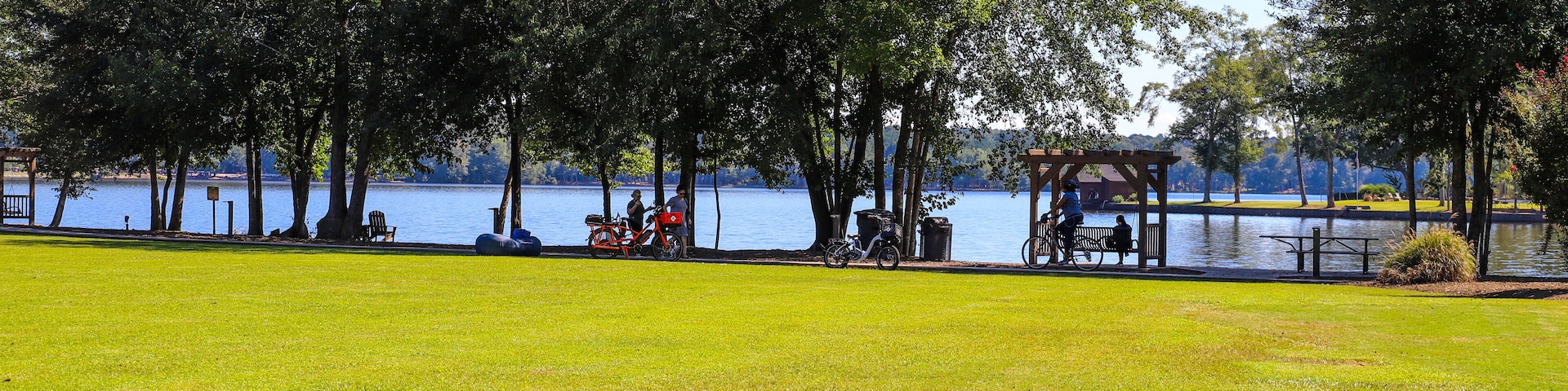Um parque com gramado, árvores, um lago e um coreto de madeira.
