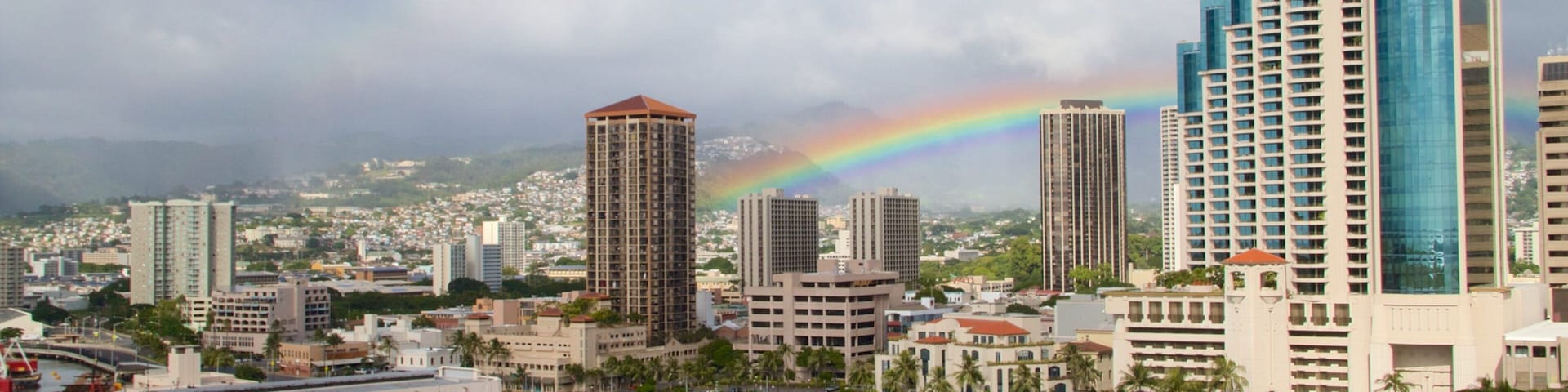 Paisagem urbana com um arco-íris marcante sobre a água, cercada por edifícios altos e uma paisagem montanhosa ao fundo.