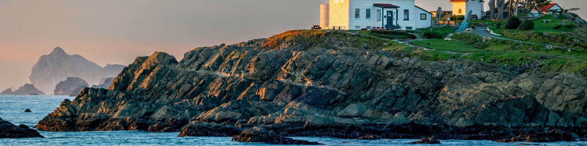 Um farol costeiro, linha costeira rochosa e montanhas ao longe.
