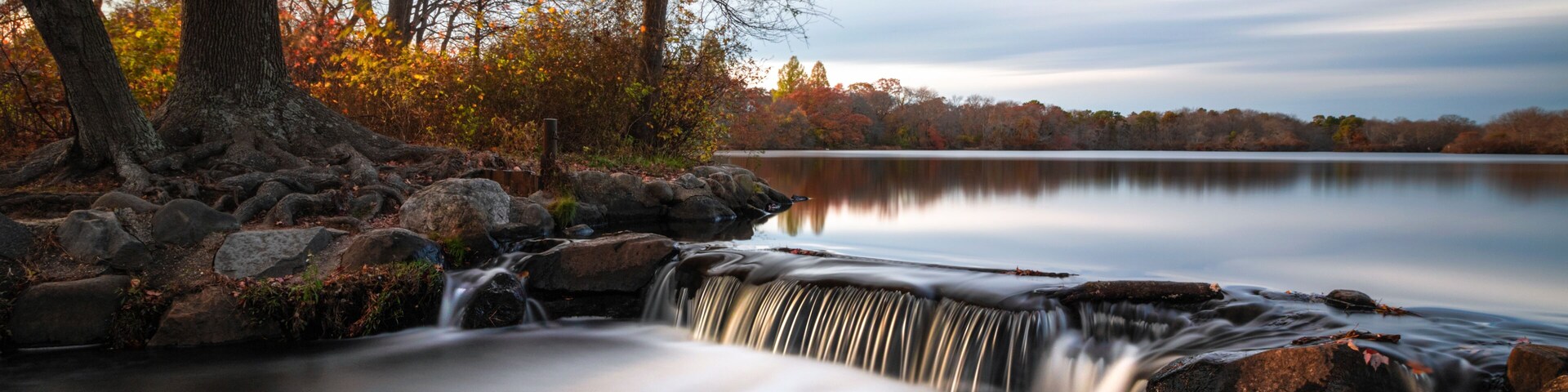 Um lago tranquilo com uma cascata, rodeado por árvores de outono e um céu claro.