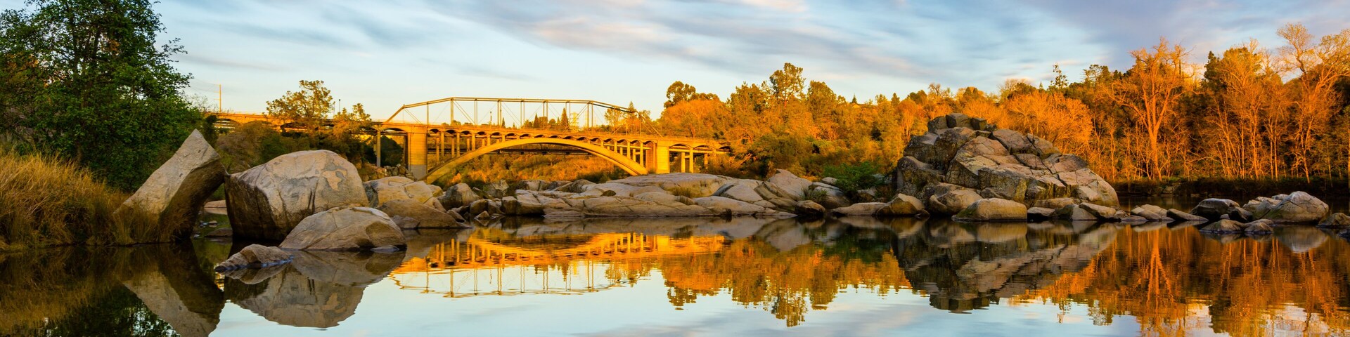 Uma ponte sobre um rio calmo, com reflexos nítidos, rodeada por folhagem outonal e grandes pedras.