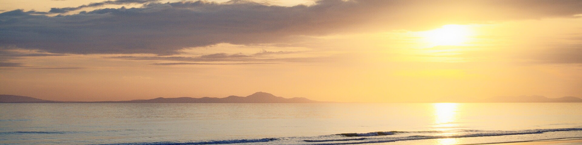 Uma praia ao pôr do sol, com duas pessoas caminhando pela costa, ondas quebrando suavemente e uma cadeia montanhosa ao longe.
