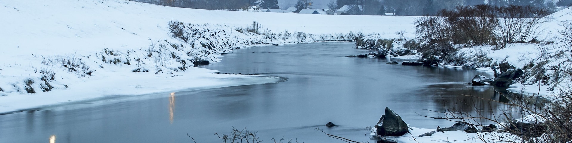 Um rio coberto de neve com um meandro, rodeado por árvores e uma cordilheira distante.