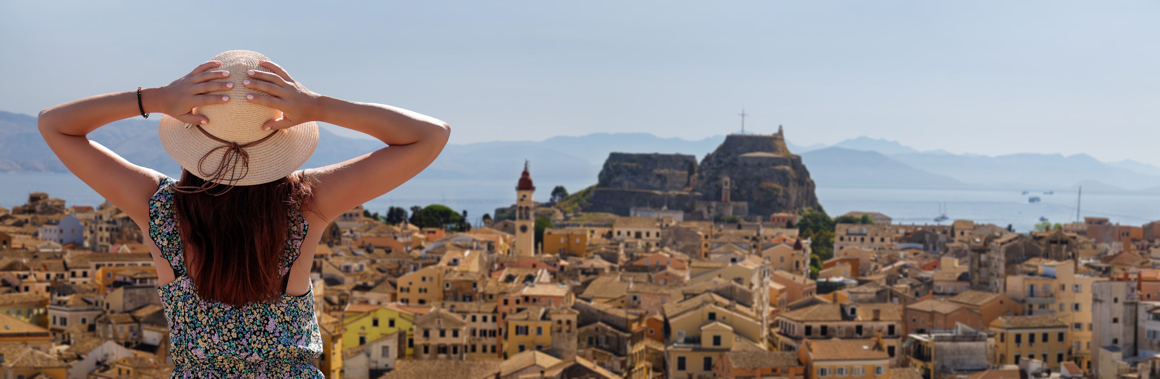 Mulher segurando um chapéu de palha, observando uma cidade histórica com uma fortaleza imponente numa colina.