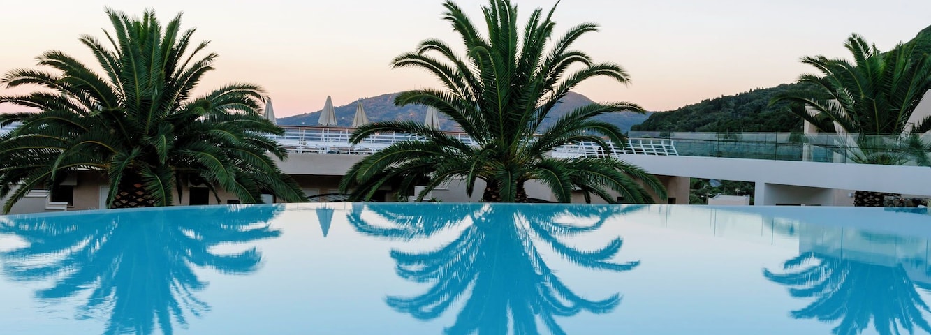 Piscina azul límpida com palmeiras e uma vista distante de um navio de cruzeiro.