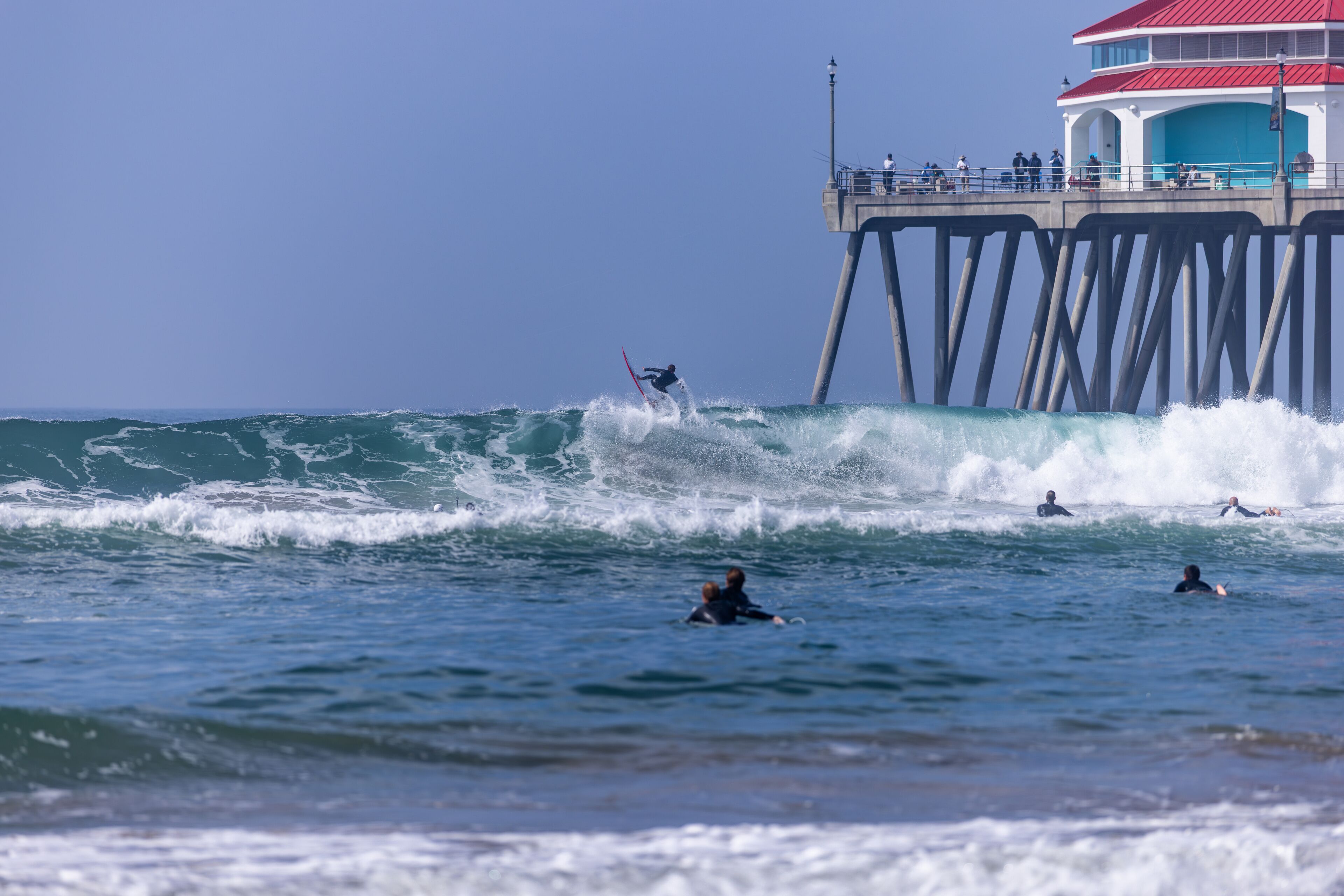 Um cais com telhado vermelho, surfistas deslizando ondas e pessoas nadando no oceano.