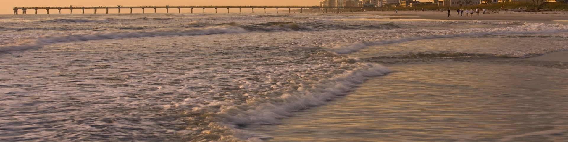 Uma praia com ondas, um cais e edifícios ao fundo.