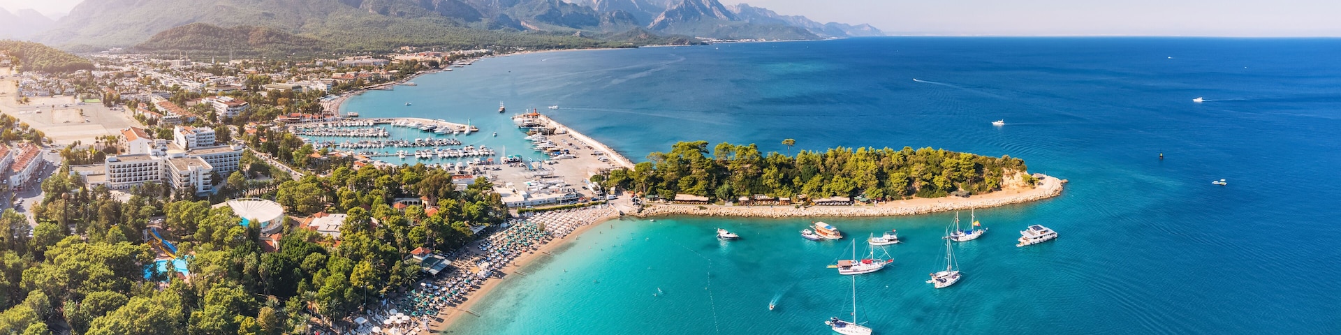 Uma cidade costeira com um porto de recreio, praia e montanhas ao fundo.