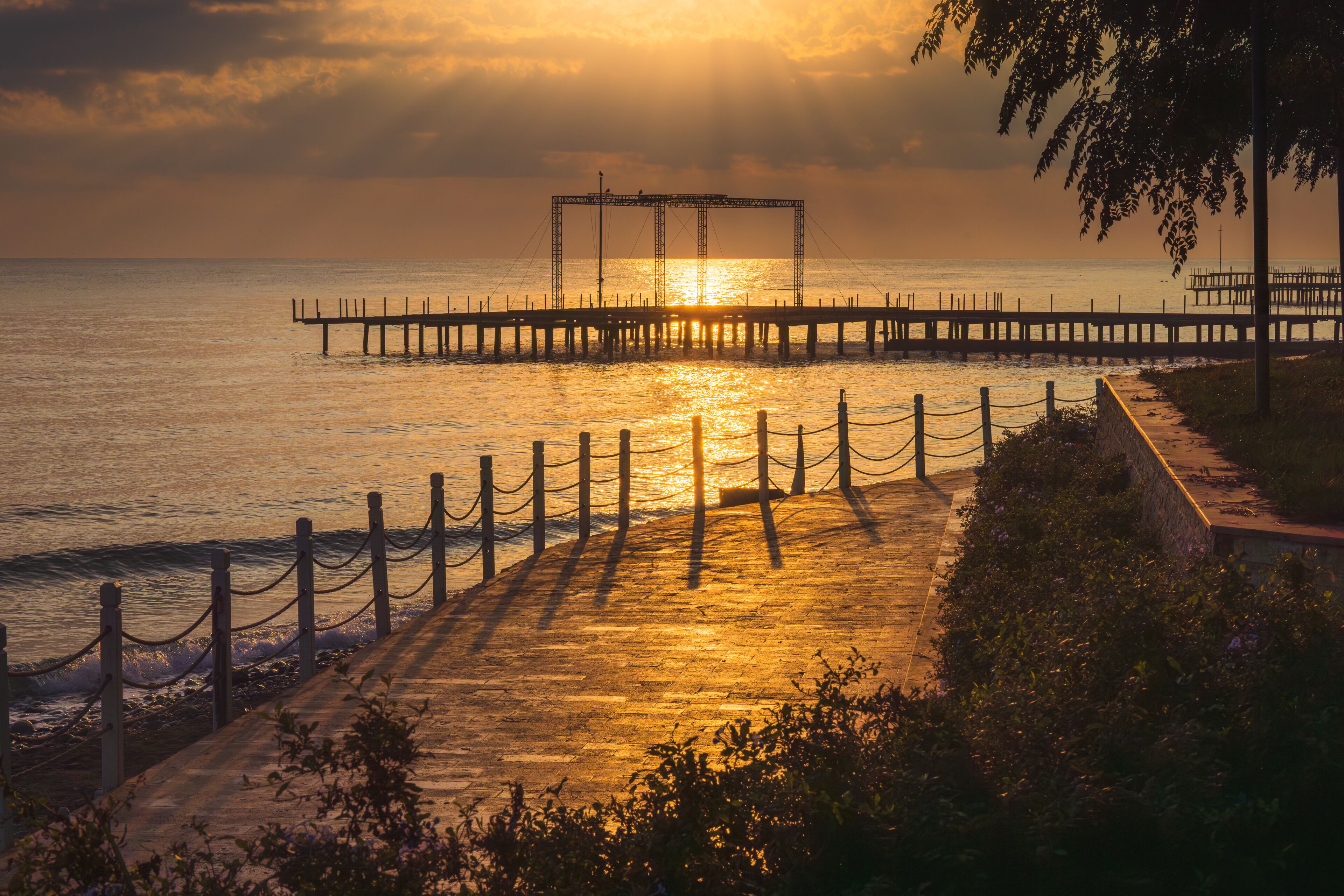 Um pôr do sol sobre um mar calmo com um cais de madeira que se estende para a água, cercado por uma cerca de arame e vegetação.