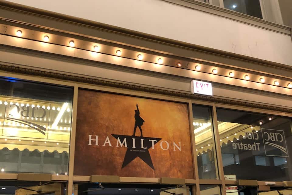 Entrada de teatro com uma marquise exibindo "Hamilton" e um letreiro de "SAÍDA" acima da porta.