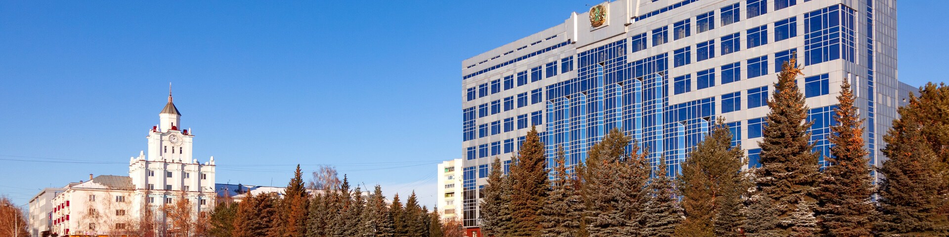 Um edifício grande e moderno com fachada de vidro, um edifício histórico com torre de relógio e uma paisagem nevada com árvores perenes.