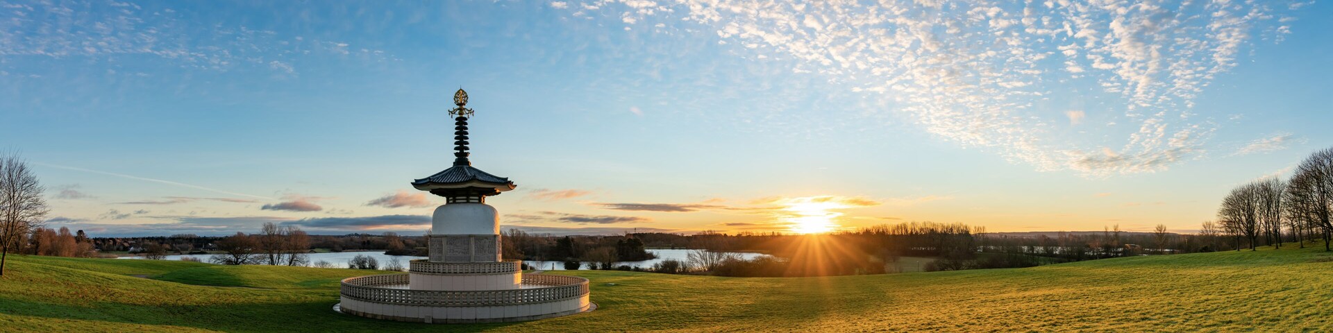 Um monumento em estilo pagoda em um campo gramado, com um lago e um pôr do sol ao fundo.