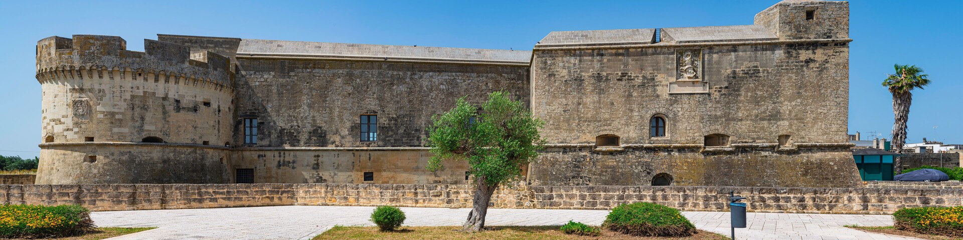 Uma fortaleza histórica de pedra com uma torre central e ameias, rodeada por um pátio pavimentado com uma palmeira e pequenos arbustos.