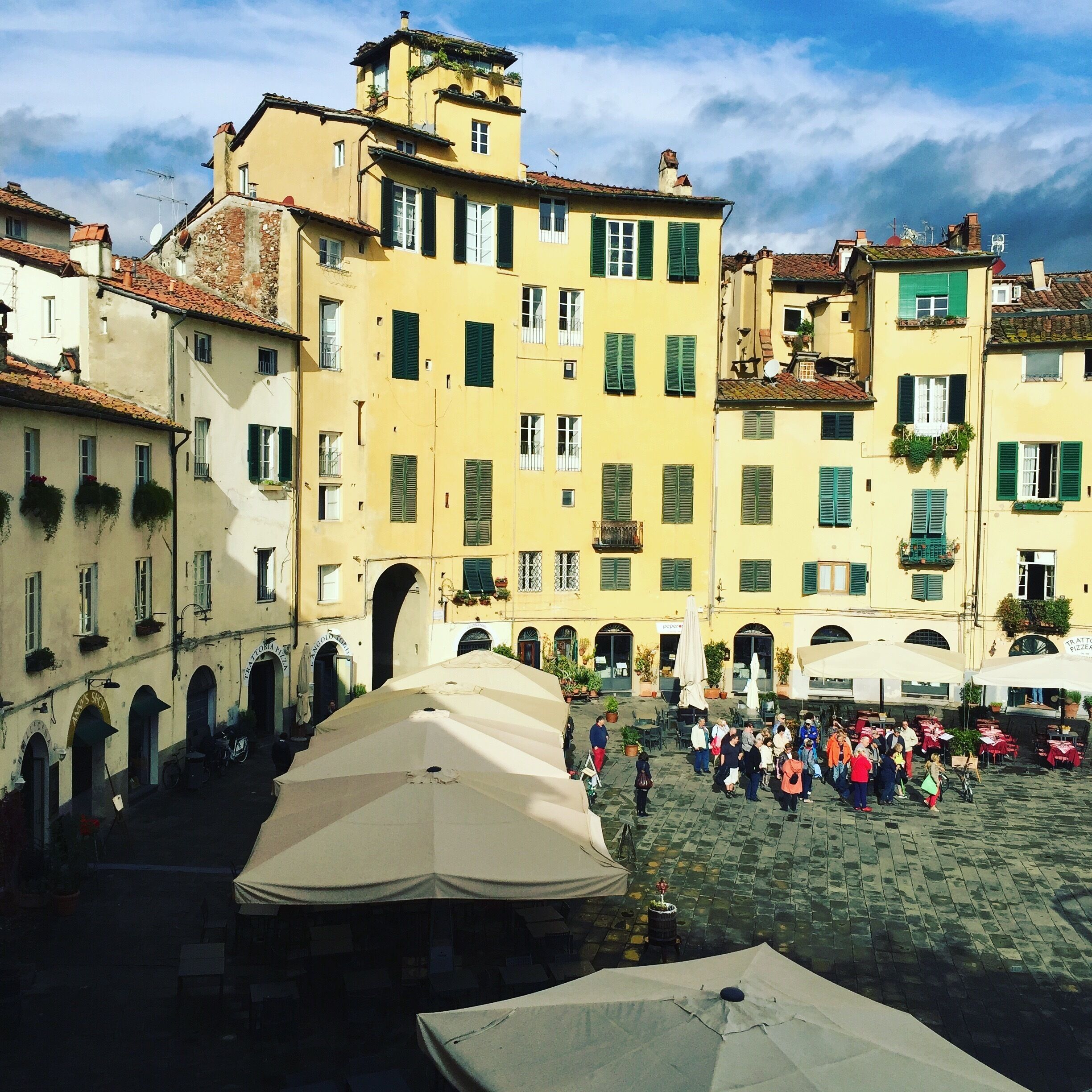 Uma praça movimentada com edifícios amarelos, venezianas verdes e áreas de estar ao ar livre.