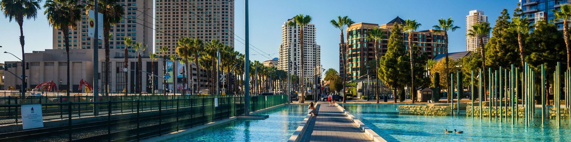 Paisagem urbana com uma piscina comprida e estreita, ladeada por palmeiras e edifícios modernos e altos.