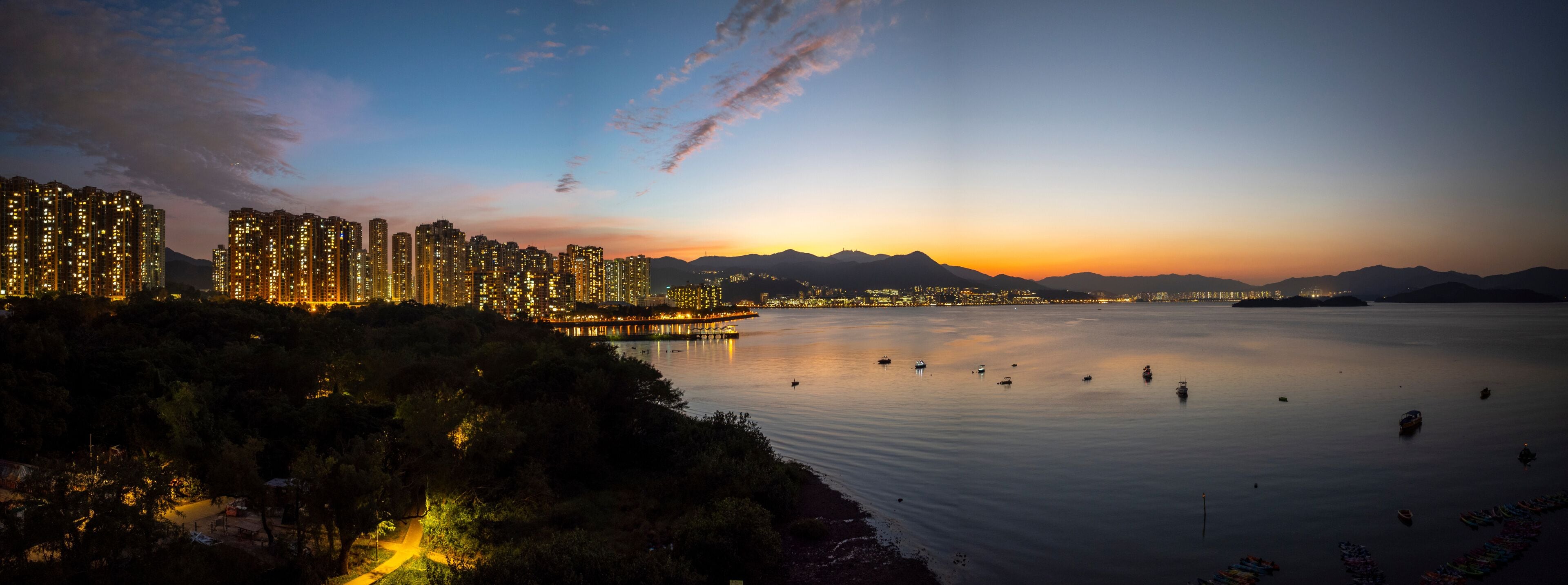 Silhueta urbana ao entardecer, com edifícios iluminados, uma superfície aquática calma e montanhas ao longe.