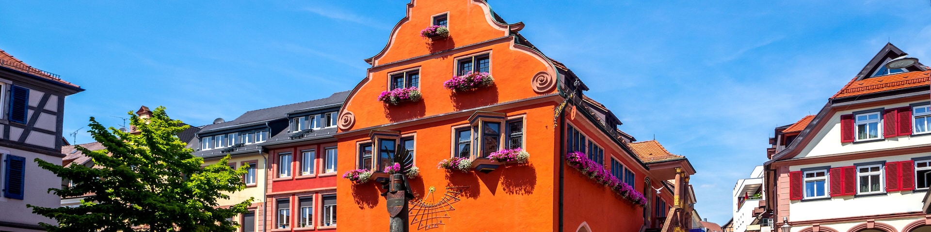 Um edifício vibrante em laranja com um pináculo verde, rodeado por arquitetura europeia tradicional.