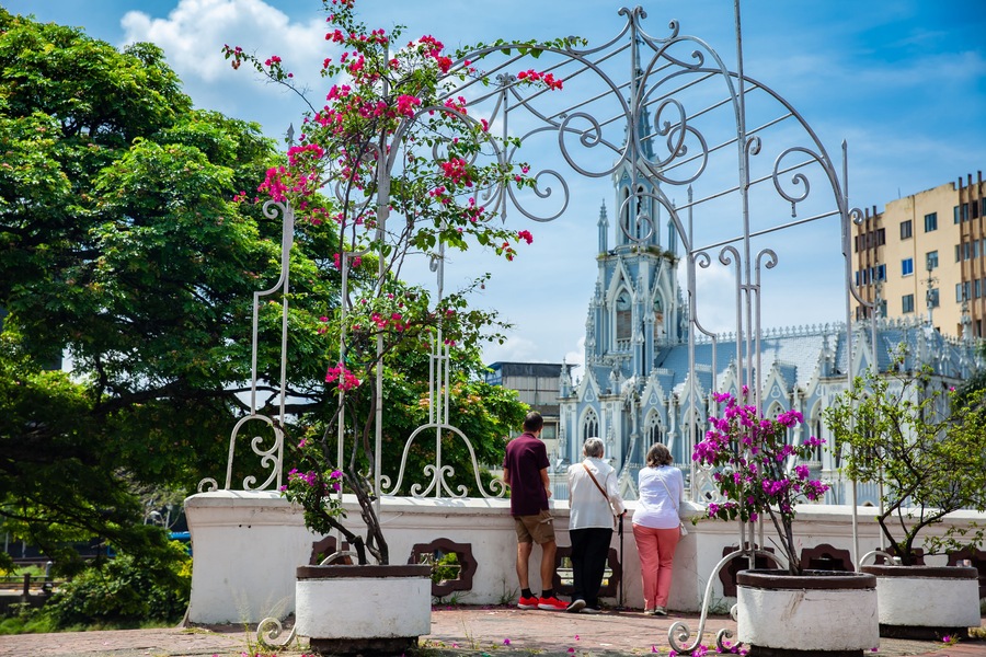 Um portão de ferro forjado com flores de buganvília rosa, uma catedral com arquitetura azul e branca e um caminho pavimentado.