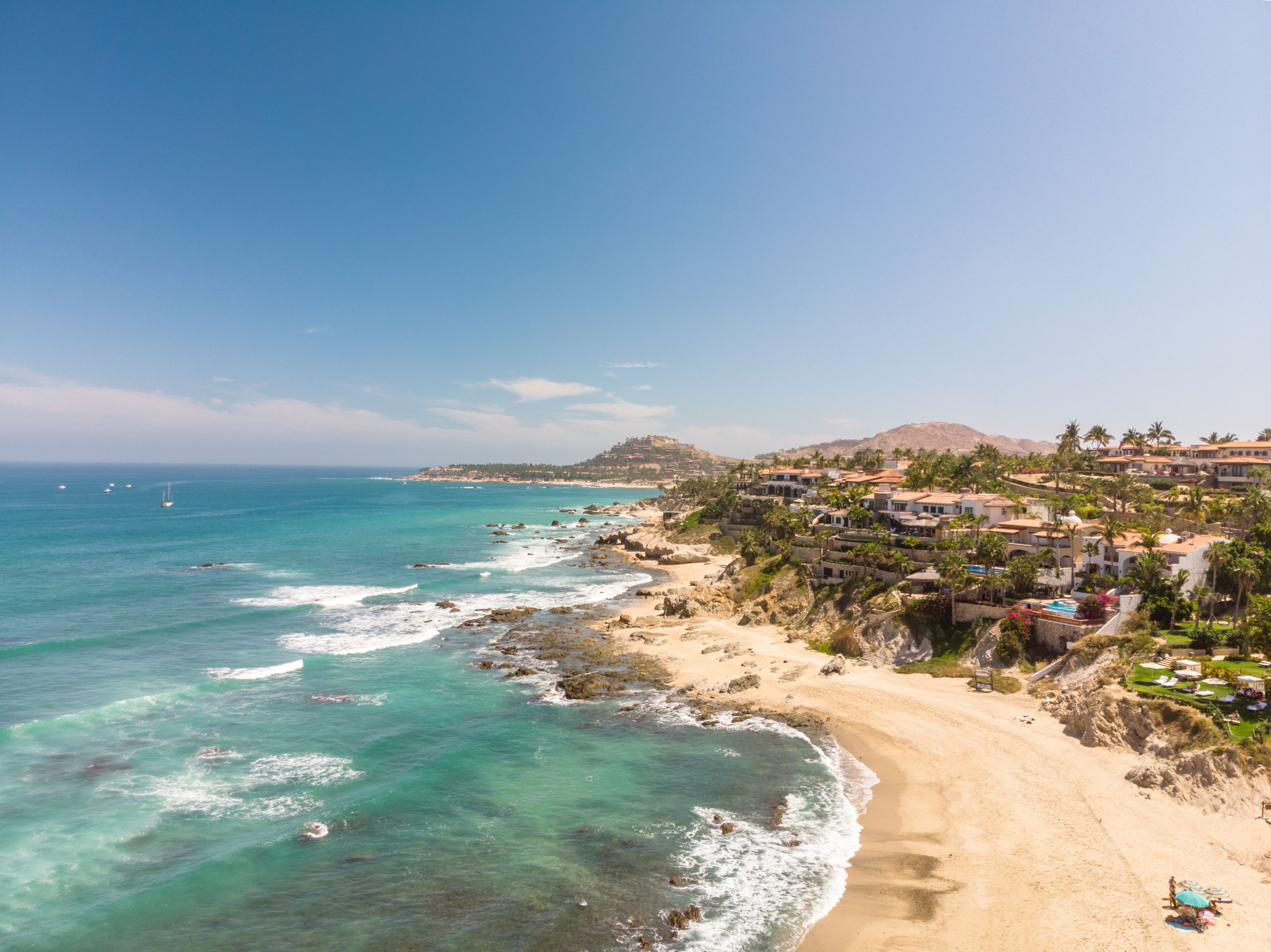 Uma vila costeira com praia, oceano e colinas ao fundo.