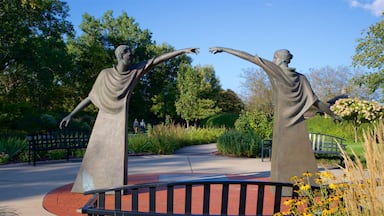Duas estátuas de bronze de figuras com braços estendidos em um parque com um caminho de tijolos e vegetação.