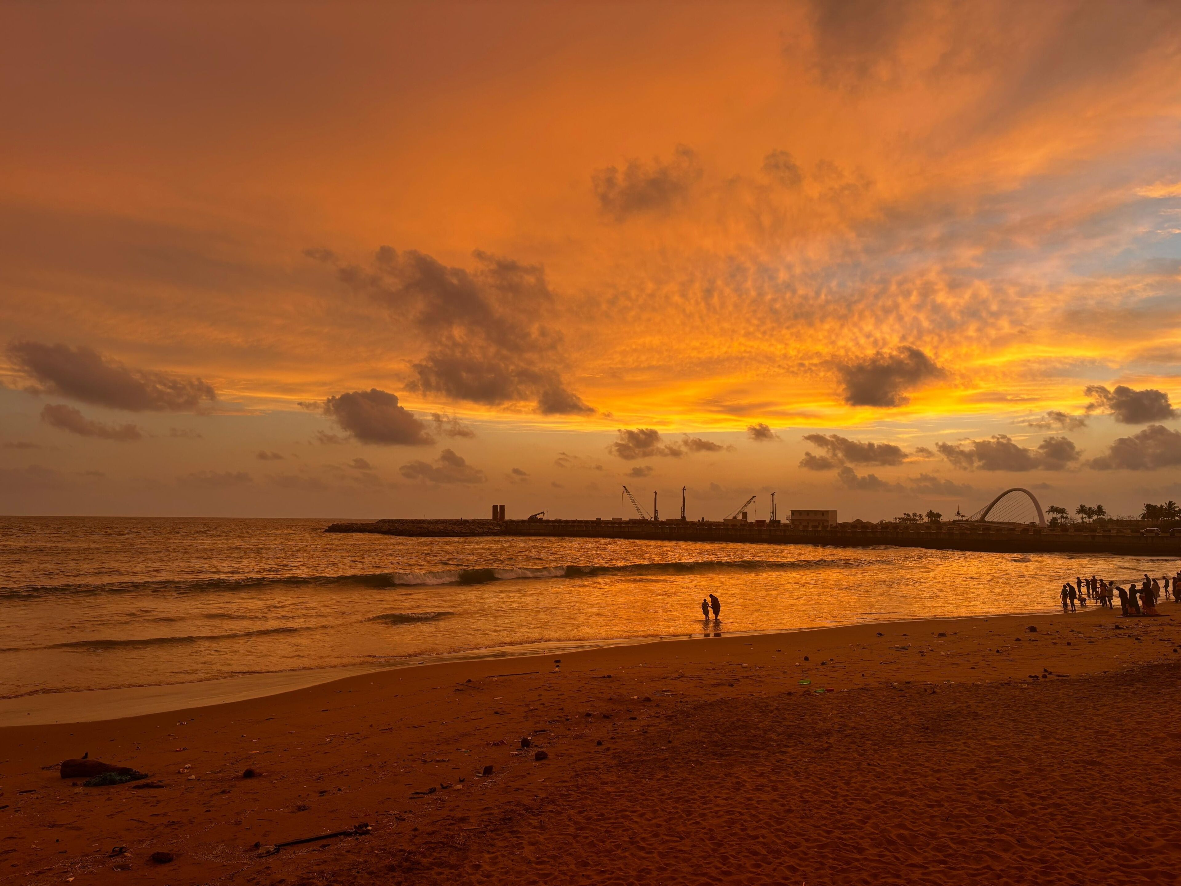 Uma praia ao pôr do sol com uma ponte curva e estruturas industriais ao fundo.