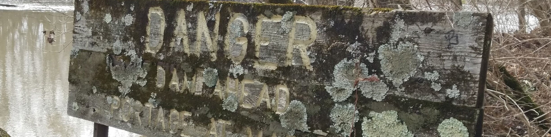 An old wooden sign warning about the lowhead dam just down stream. There are newer looking metal signs. The lichen encrusted sign reads "Danger, Dam Ahead. Portage at Marker". This can be found east of Antrim lake on the western shore of the river, along the trails that snake through the wooded area of the park. #urbanjungle