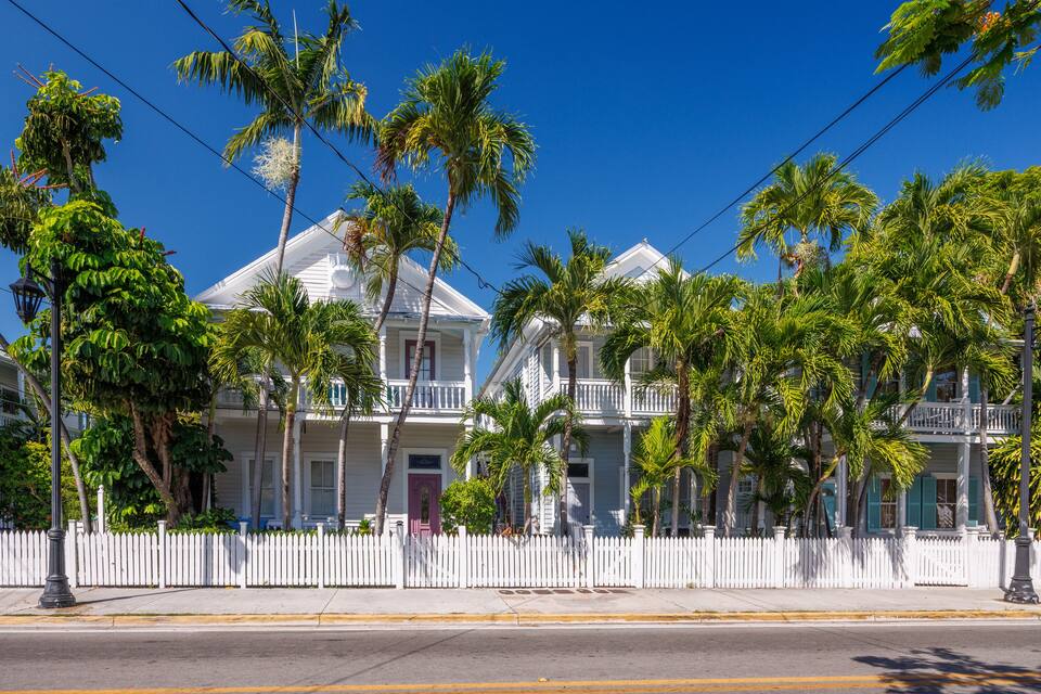 Um edifício branco em estilo colonial com cerca branca de piquete, cercado por palmeiras.