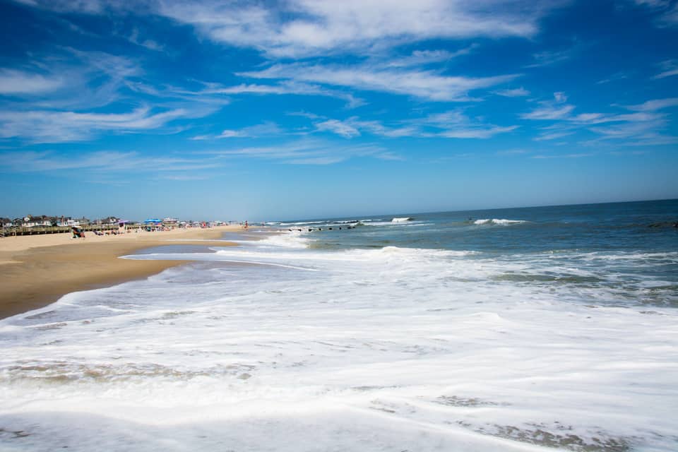 Uma praia com ondas, céu claro e um litoral distante com edifícios.