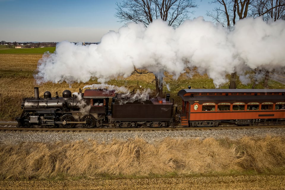 Uma locomotiva a vapor puxando um vagão de passageiros por uma paisagem rural com campos e árvores.