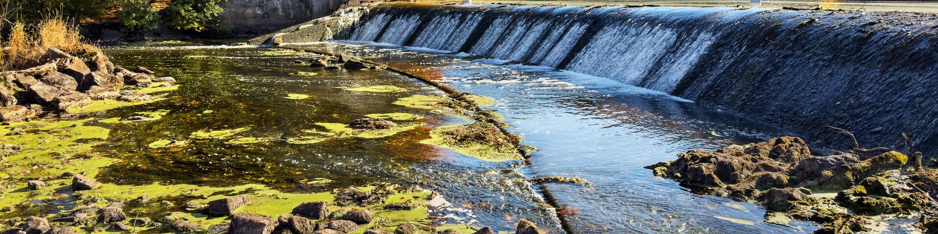 Um rio serpenteando por uma paisagem rochosa, com uma barragem e árvores em tons de outono ao fundo.