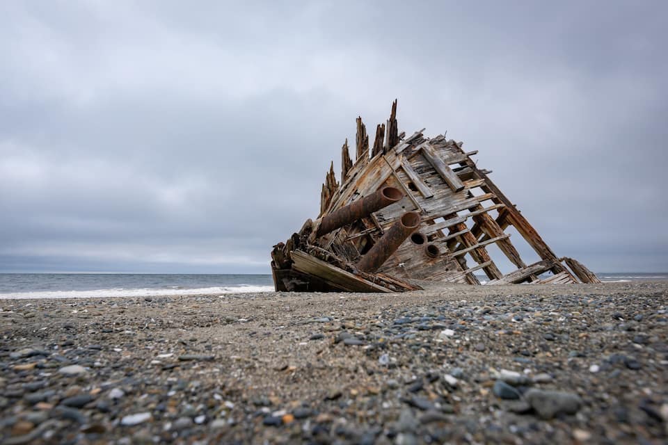 Um naufrágio abandonado numa praia de pedras com o oceano ao fundo.