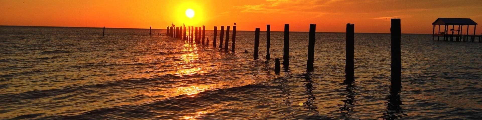 Pôr do sol sobre um mar calmo, com estacas de madeira e uma pequena estrutura à direita.