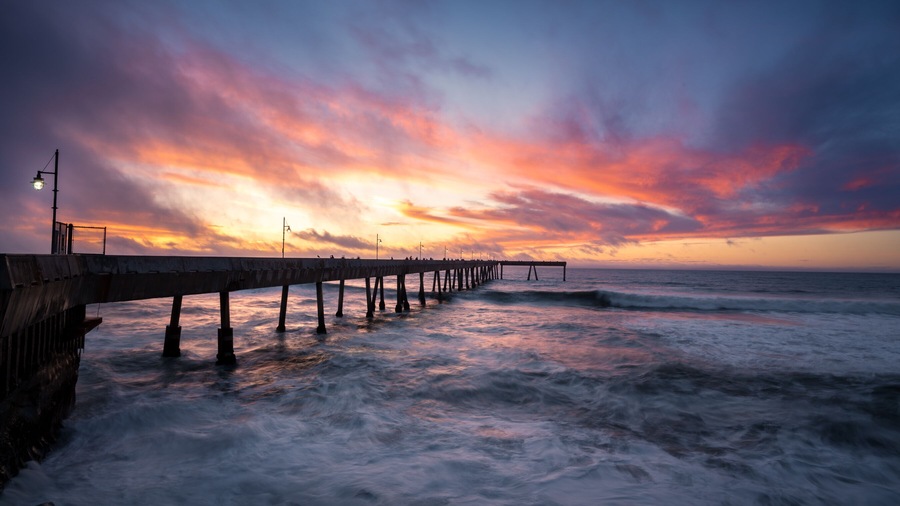 Um píer que se estende para o oceano, com o pôr do sol ao fundo.
