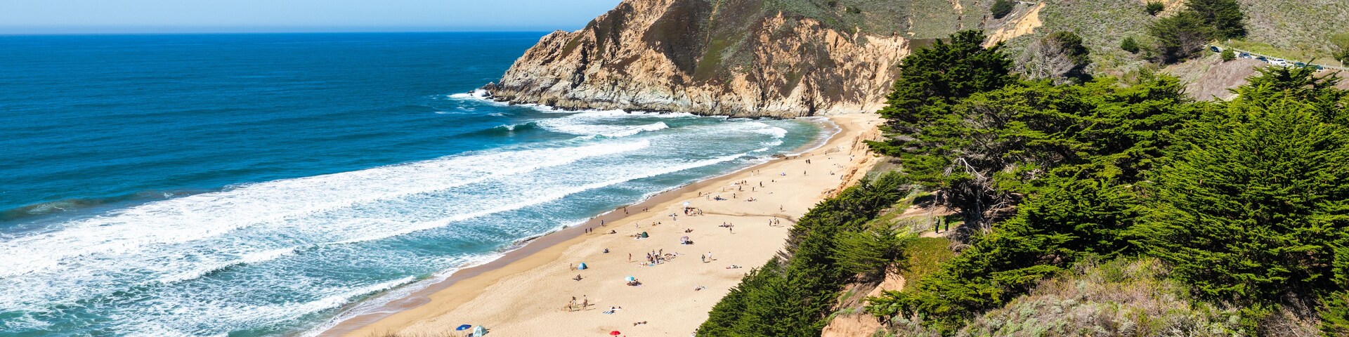 Paisagem costeira com uma praia arenosa, falésias rochosas e vegetação verde exuberante.