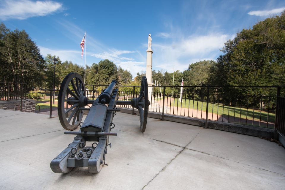 Um canhão em um parque, com uma estátua e bandeiras americanas ao fundo.