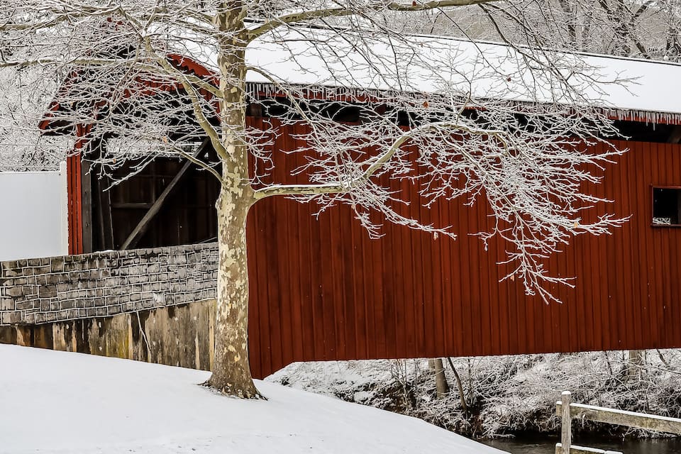 Uma ponte de madeira coberta de neve, um edifício vermelho e uma árvore com galhos nus.