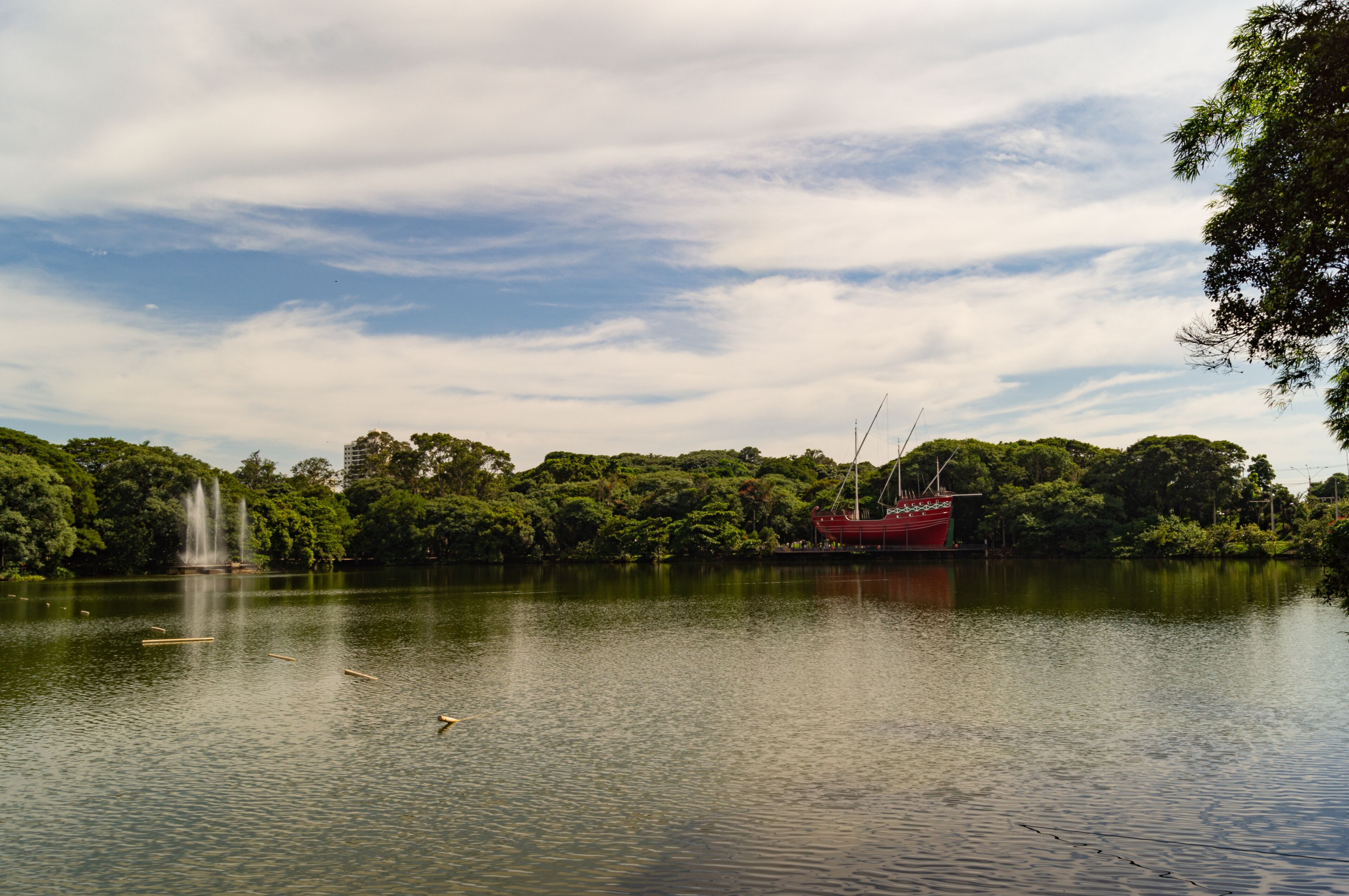 Um lago tranquilo com uma fonte, rodeado por árvores verdes densas e um barco vermelho atracado na margem.
