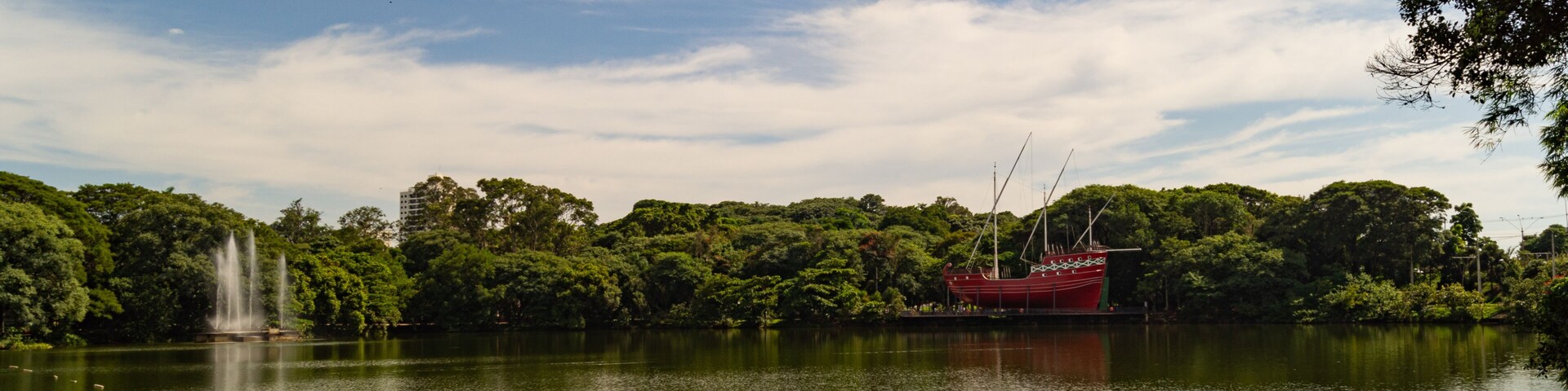 Um lago tranquilo com uma fonte, rodeado por árvores verdes densas e um barco vermelho atracado na margem.
