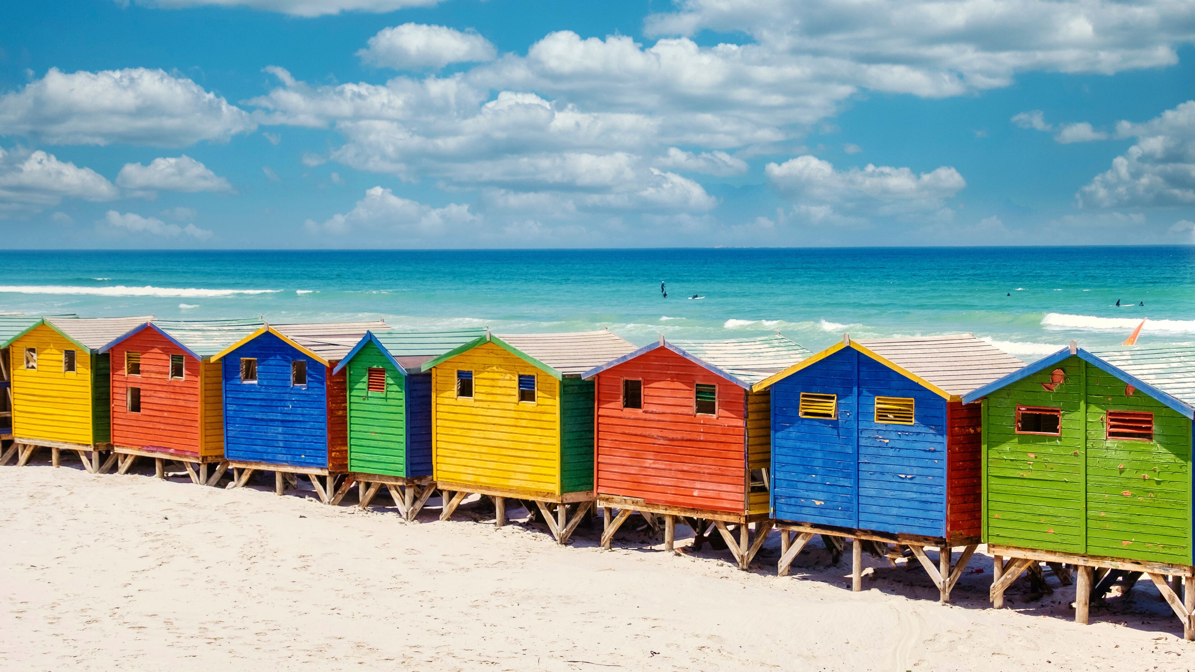 Uma fileira de cabanas coloridas sobre palafitas, ao longo de uma praia arenosa, com o oceano ao fundo.