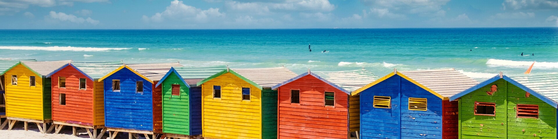 Uma fileira de cabanas coloridas sobre palafitas, ao longo de uma praia arenosa, com o oceano ao fundo.