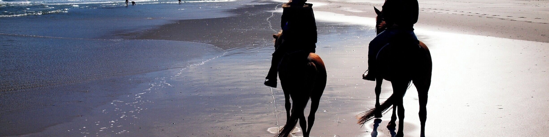 Duas pessoas cavalgam a cavalo numa praia, com montanhas ao fundo.