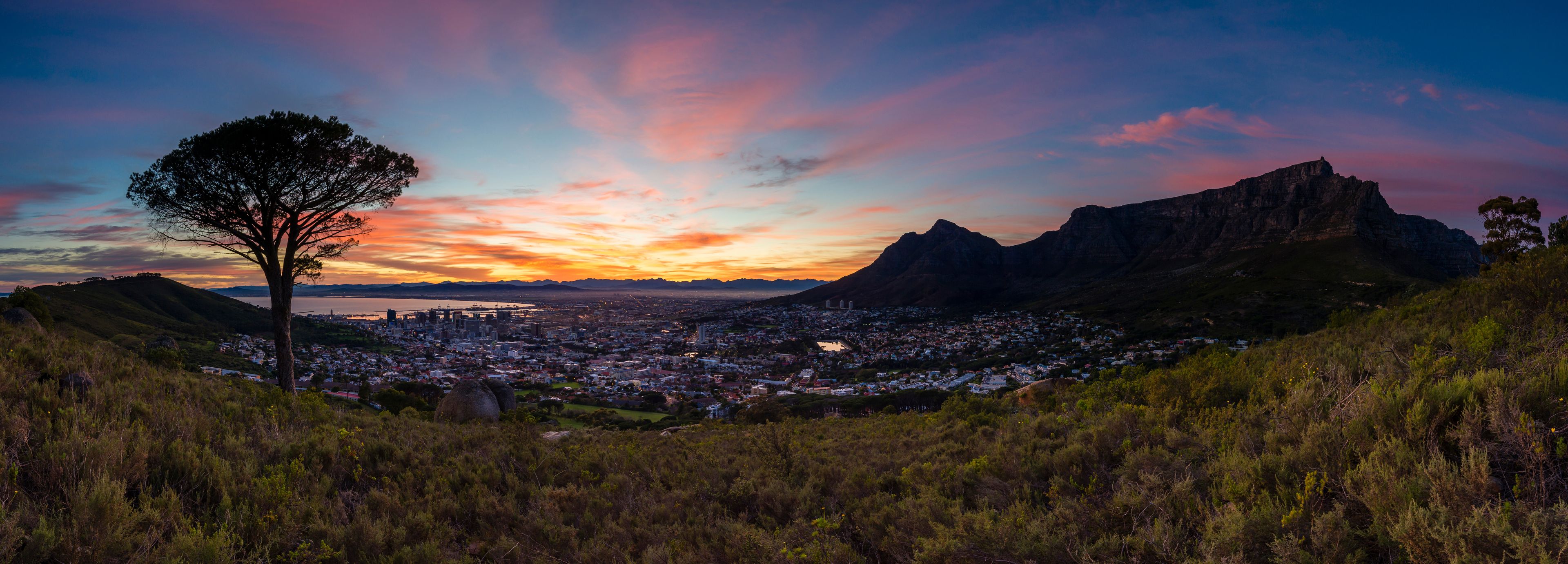 Paisagem urbana ao pôr do sol, com uma cadeia de montanhas imponente ao fundo.