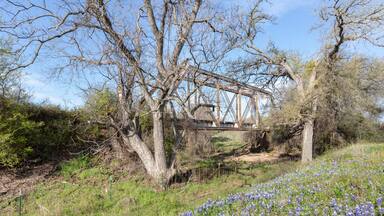 Uma ponte enferrujada sobre um riacho, rodeada por árvores e flores silvestres.