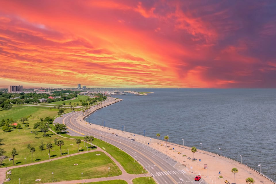 Paisagem costeira urbana com uma estrada sinuosa, áreas verdes de parque e um pôr do sol sobre o oceano.